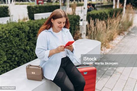 도시 배경에 스마트 폰을 사용하여 빨간 여행 가방을 가진 젊은 빨간 머리 여자 야외 현대 벤치에 앉아있는 행복한 소녀 도시 생활 방식 개념 기술 배너 여자에 대한 스톡 사진