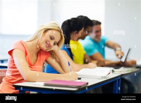 Beautiful Female Student Writing Notes In Classroom With Classmates On