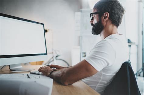 잘생긴 수염된 전문 건축가 현대 로프트 스튜디오데스크톱 컴퓨터와 사무실에서 일 하는 안경 착용 화이트 빈 화면을 표시 합니다 흐리게 배경입니다 수평 건축가에 대한 스톡 사진