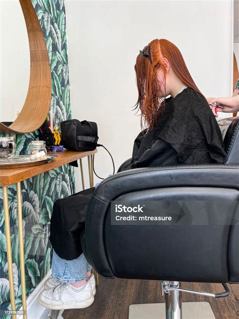 검은 이발사의 망토를 입고 미용실 가죽 안락의자에 앉아 있는 매력적인 빨간 머리 여자의 이미지 젖은 머리를 자르는 알아볼 수 없는 사람 머리를 자르는 플라스틱 머리핀