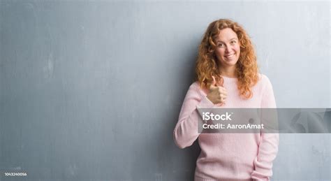 젊은 빨강 머리 여자 회색 그런 지 벽 입고 위에 분홍색 스웨터 행복 엄지손가락 손으로 제스처를 하 고 승인 성공을 보여주는 카메라를 보고 하는 식 곱슬 머리에 대한 스톡