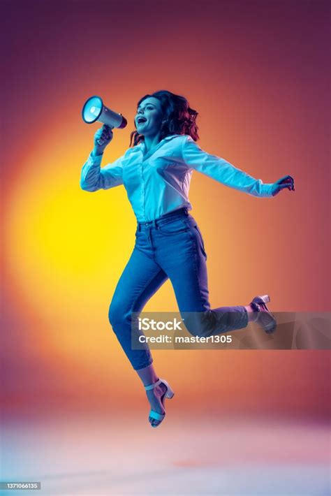 네온 빛 필터에 오렌지 배경에 고립 된 긴 곱슬 머리 점프 와 행복한 젊은 아름다운 소녀의 전체 길이 초상화 감정 아름다움 패션의 개념 휴대용 마이크에 대한 스톡 사진 및