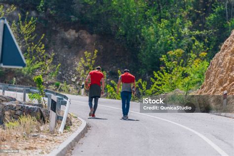 오르막길을 오르고 내리막 길을 준비하는 동안 두 명의 남성 스케이트보더가 롱보드를 손에 들고 있었다 빨간 티셔츠 녹색 모자 슈퍼 쿨 선글라스를 착용 20 24세에 대한 스톡
