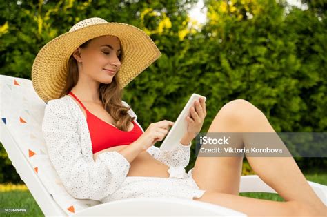 빨간 수영복과 모자를 쓴 예쁜 여자는 디지털 태블릿을 사용하고 긴 의자에서 일광욕을 하면서 미소를 짓는다 30 34세에 대한 스톡 사진 및 기타 이미지 Istock