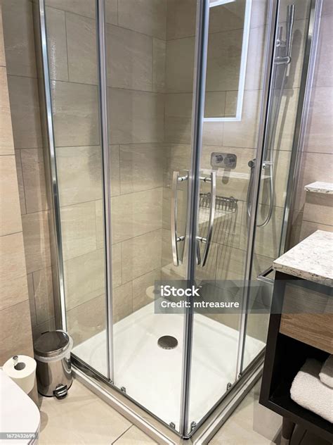 흰색 욕실 스위트 별도의 유리 샤워 부스 흰색 접힌 수건 욕실 세면대 카운터 아래 나무 선반 유닛 화장실 롤 홀더 베이지색 석회화 벽 및 바닥 타일 전경에 초점 0명에 대한