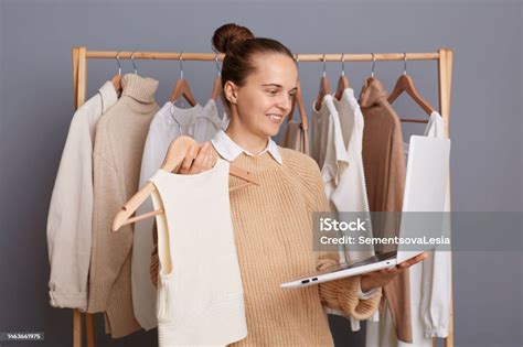 베이지색 스웨터를 입고 회색 벽에 서서 옷을 입은 웃는 여성 스타일리스트 블로거의 초상화가 선반의 옷장에 걸려 있어 Vlog 콘텐츠를 만들고 구독자에게 세련된 의상을 보여