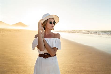 하얀 드레스를 입고 아름다운 모래 해변에서 포즈를 취하는 여름 모자를 쓴 패셔너블한 여성 푸에르테벤투라에 대한 스톡 사진 및 기타 이미지 푸에르테벤투라 개념 관광객