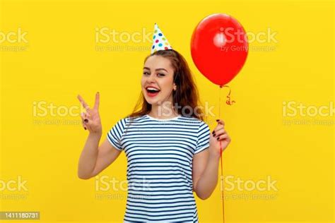 생일 모자를 쓴 예쁜 젊은 여성은 노란색 배경에 빨간색 팽창 헬륨 풍선을 들고 평화 제스처를 취합니다 휴일과 파티 개념 20 24세에 대한 스톡 사진 및 기타 이미지
