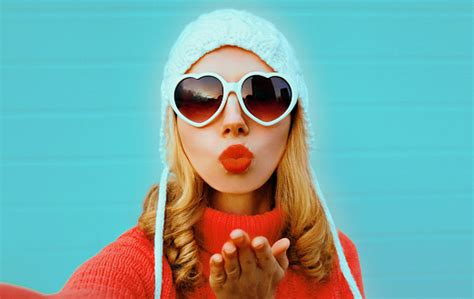 빨간 니트 스웨터 흰색 모자 파란색 벽 배경에 하트 모양선글라스를 입고 셀카를 찍기 위해 손을 뻗는 아름다운 젊은 여성의 겨울 초상화 선글라스에 대한 스톡 사진 및 기타