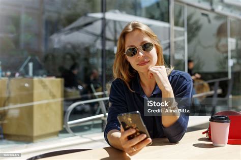 술 한 잔을 든 예쁜 힙스터 소녀가 길거리 여름 카페 테이블에 앉아 있다 커피 브레이크를 가져 가라 거리에 대한 스톡 사진 및 기타 이미지 Istock