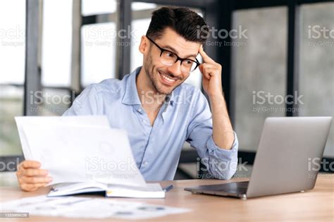 행복한 성공적인 자신감 젊은 백인 사업가 매니저 또는 프리랜서는 이익을 즐기고 비즈니스 문서를 들고 노트북 화면을보고 미소 사무실에서 테이블에 앉아 안경을 착용 30 39