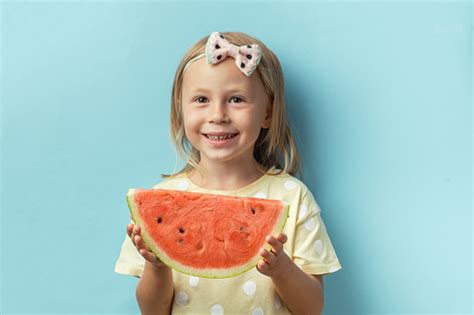 파란색 배경에 육즙이 많은 붉은 수박의 큰 조각을 즐기는 귀여운 소녀 노란색 티셔츠에 금발 머리를 가진 행복한 백인 소녀의 초상화 더운 여름 개념 2 3 살에 대한 스톡