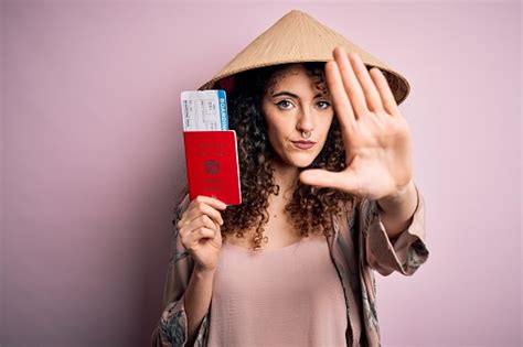 곱슬 머리와 피어싱 아시아 모자를 입고 일본 여권을 들고 있는 아름다운 관광 여성은 진지하고 자신감있는 표정으로 정지 표지판을하고 방어 제스처 경계 표지에 대한 스톡 사진