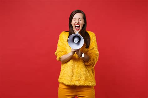 미친 젊은 여자 눈 노란색 모피 스웨터에 스튜디오에서 밝은 빨간 벽 배경에 고립 된 확성기에 비명 사람들이 진심으 감정 라이프 스타일 개념 복사 공간을 비웃는 다 가을에