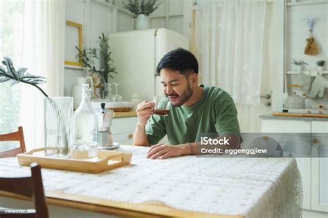 녹색 티셔츠를 입은 젊은 아시아계 일본인 남자가 식탁에서 커피 한 잔을 즐기고 있고 수염을 기른 젊은 남자는 눈을 감고 모닝 커피의 풍부한 향기와 부드러운 맛을 음미하고