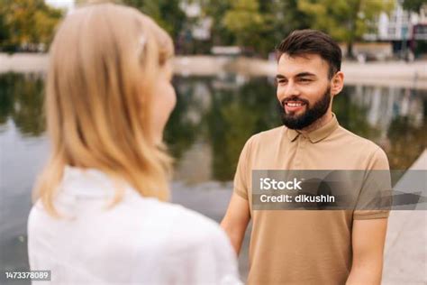 쾌활한 수염을 기른 남자가 도시 공원의 아름다운 호수 옆에서 여자 친구와 손을 잡고 서서 이야기하고 서로를 바라보고 있습니다 20 24세에 대한 스톡 사진 및 기타 이미지