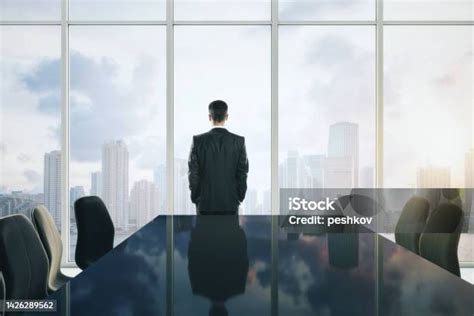 현대적인 회의실 내부에 서있는 젊은 사업가의 뒷모습은 테이블 의자 및 하늘 구름 전망과 일광이있는 탁 트인 창문에 반사되어 있습니다 Ceo 및 컨퍼런스 개념 회의실에 대한