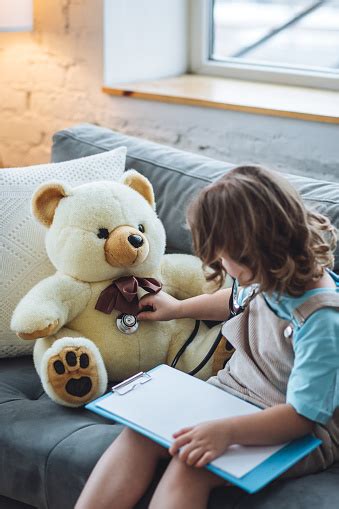 귀여운 어린 소녀가 집에서 큰 푹신한 테디 베어와 함께 의사가 되어 소파에 앉아 청진기로 심장 박동을 확인합니다 아늑한 조명 4
