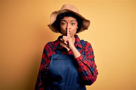 앞치마와 모자를 쓰고 곱슬 머리를 한 젊은 아프리카 계 미국인 아프로 농부 여성은 입술에 손가락으로 조용히 해달라고 부탁합니다 침묵과 비밀 개념 농부에 대한 스톡 사진 및