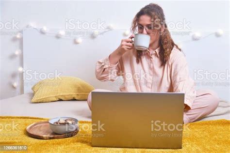 귀여운 여자는 홈 침대에서 커피를 마십니다 격리로 인해 격리 중에 원격으로 작동하는 컴퓨터와 빨간 머리 소녀 일함에 대한 스톡