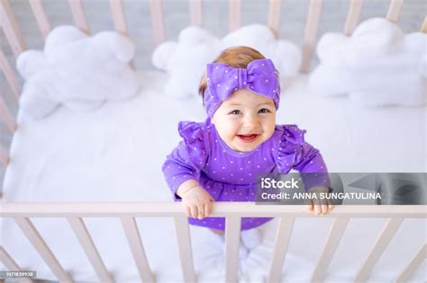 유아용 침대에 귀여운 아기 소녀는 라일락 옷을 입고 옆으로 잡고 있고 머리에 활을 얹고 있다 재미 있은 작은 아기 평면도 어린이 용품의 개념 6 11 개월에 대한 스톡 사진