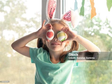 토끼 귀를 가진 어린 소녀가 창턱에 앉아서 부활절 달걀을 눈에 칠하고 얼굴을 만들고 웃고 있습니다 2명에 대한 스톡 사진 및 기타 이미지 Istock