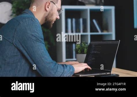 Man In Denim Shirt Coding Software At Laptop Desk With Computer