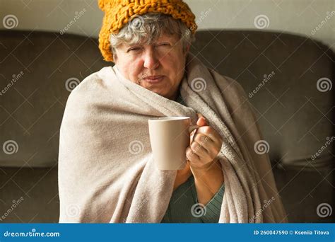 Elderly Woman In Ginger Cap Wrapped In Blanket And Drinking Cup Of Tea