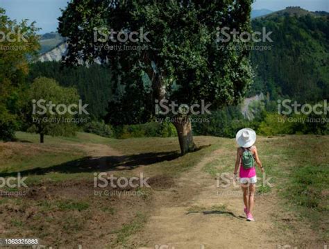 흰 모자와 배낭을 입은 한 여성 등산객이 길을 따라 숲속으로 걸어들어간다 그녀의 옆에 큰 나무와 배경에 산 50 54세에 대한 스톡 사진 및 기타 이미지 Istock