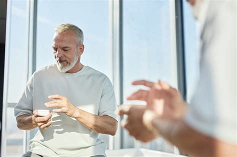 크림 항아리를 들고 있는 성숙한 남자는 실내에서 자기 관리 루틴 중 피부를 보습합니다 55 59세에 대한 스톡 사진 및 기타 이미지 55 59세 가정 생활 개성 개념