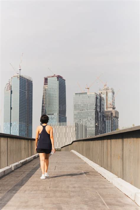 Woman Exercise Walking On The Bridge Hand Holding Bottle Water With City Building View Stock