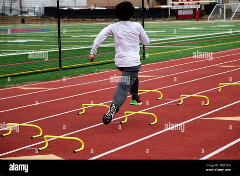 Rear View Of A High School Boy Running The Wicket Drill On A Red Track Over Yellow Mini Hurdles