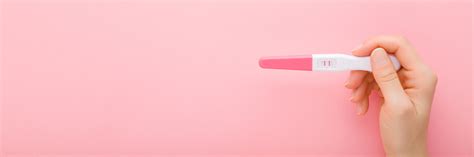 젊은 성인 여성 손 밝은 분홍색 테이블 배경에 두 줄무늬와 임신 테스트를 들고 파스텔 색상 긍정적 인 결과 근접 촬영 관점 샷