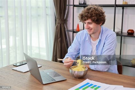 젊은 백인 곱슬머리 남자는 테이블에 앉아 젓가락과 컴퓨터로 작업실에서 점심으로 맛있는 라면을 먹는다 건강에 해롭고 값싼 음식 라이프 스타일 개념 25 29세에 대한 스톡