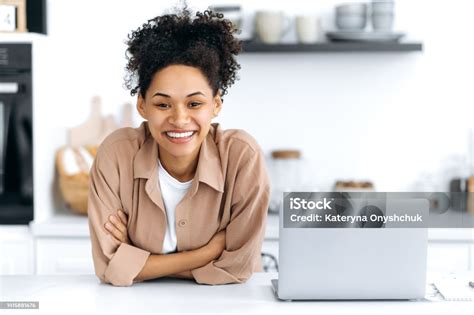 초상화 사랑스러운 긍정적 인 곱슬 머리 아프리카 계 미국인 젊은 여성 캐주얼 한 세련된 옷 여학생 또는 프리랜서 노트북 근처의 부엌에서 집에 서서 카메라를보고 행복하게 미소