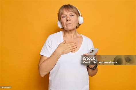 나이 든 여성이 헤드폰을 끼고 선명한 노란색 배경 앞에 서서 음악을 듣고 있습니다 그녀는 스마트폰을 들고 마음을 어루만지며 음악에 대한 깊은 정서적 연결을 보여줍니다 55