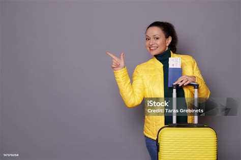 노란색 재킷을 입은 임산부 비행기 표와 여행 가방을 들고 포즈를 취하고 미소를 지으며 회색 배경의 복사 공간을 가리키고 있다