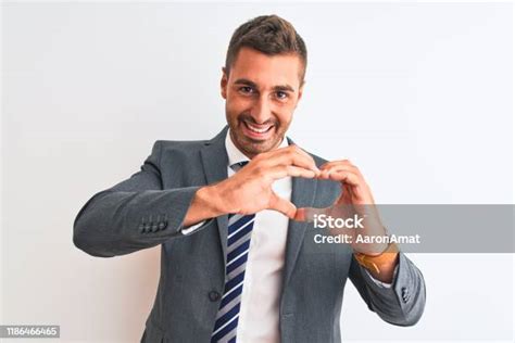 양복을 입고 고립 된 배경 위에 넥타이를 매고 사랑으로 미소 짓는 젊은 잘생긴 사업가는 손으로 심장 기호 모양을하고 있습니다 로맨틱 한 개념 하트 모양에 대한 스톡 사진 및