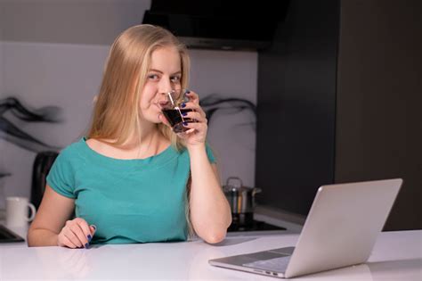 녹색 티셔츠를 입은 예쁜 금발 소녀는 갈색 액체 음료와 함께 유리를 들고 회색 방에서 노트북 근처에서 일하거나 공부합니다 커피 차 알코올 위스키 주스 T 셔츠에 대한 스톡