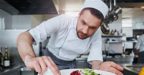 흰 유니폼을 입은 검은 머리의 백인 요리사가 샐러드 요리를 마칩니다 그는 접시를 더 잘 보이게하기 위해 접시의 측면을 닦습니다 사진 Unsplash의 레스토랑 주방 이미지