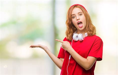 헤드폰와 깜짝 놀라게 하 고 손으로 제시 하 고 손가락으로 가리키는 동안 카메라에 웃 고 모자 고립 된 배경 위에 젊은 아름 다운 Hipster 여자 가리키기에 대한 스톡