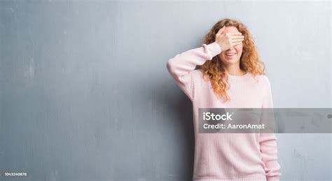 분홍색 스웨터 웃 고 손으로 덮고 눈 깜짝 하는 얼굴에 웃음을 입고 회색 그런 지 벽에 빨강 머리 젊은 여자 눈 먼 개념입니다 가리기에 대한 스톡 사진 및 기타 이미지