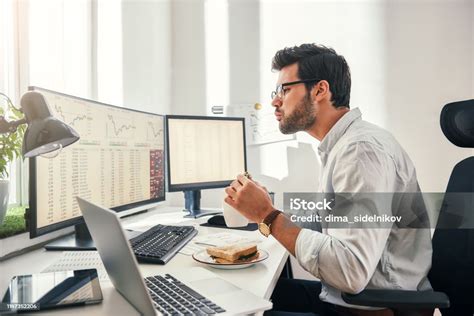 배고픈 사업가 젊은 수염 상인의 측면도 그의 현대 사무실에서 금융 데이터와 모니터 화면을 보면서 신선한 샌드위치를 먹고 있다 샌드위치 음식에 대한 스톡 사진 및 기타 이미지