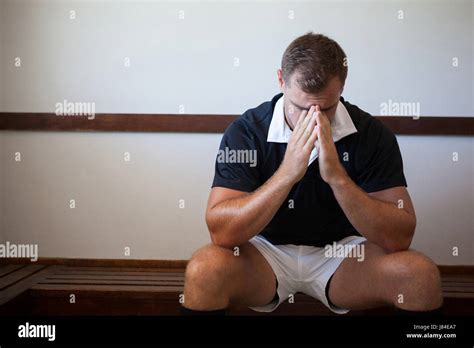 Thoughtful Rugby Player Sitting On Bench Against White Wall In Locker