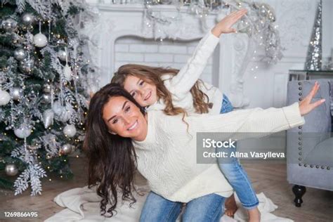 새해 전야에 행복한 가족 젊은 아름다운 어머니와 흰색 아늑한 스웨터와 담요에 작은 귀여운 딸은 장식 된 크리스마스 트리 아래에서 재미와 포옹을합니다 축제 홈 클래식 인테리어