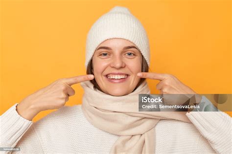 흰색 스웨터 모자 스카프를 착용하고 검지 손가락으로 이빨 미소를 가리키며 미백이나 치과 의사 방문 후 잘 관리된 하얀 치아를 보여주는 행복한 여자 노란색 배경 위에 고립되어