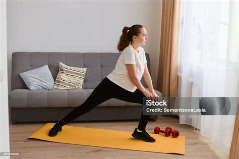 흰색 티셔츠와 검은 색 레긴을 입고 집에서 스포츠 운동을하고 다리를 스트레칭하고 훈련 전에 워밍업을하는 포니 테일과 스포티 한 매력적인 여성의 실내 샷 30 34세에 대한