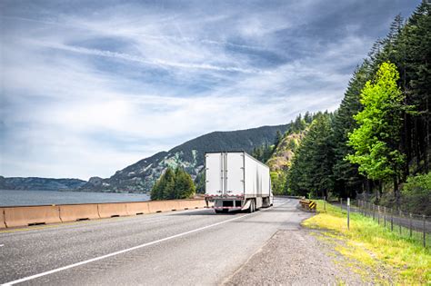 컬럼비아 협곡 지역의 강을 따라 구불구불한 도로에서 달리는 드라이 밴 세미 트레일러에서 상업용 화물을 운반하는 대형 장비 흰색 세미 트럭 트랙터 0명에 대한 스톡 사진 및