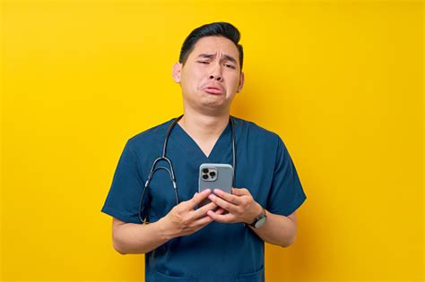 슬픔에 잠긴 젊은 아시아 남자 의사나 간호사는 파란 유니폼을 입고 휴대폰을 들고 노란 배경에 고립된 카메라를 들고 있다 의료 의학 개념 30 34세에 대한 스톡 사진 및