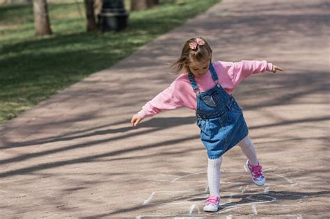 분홍색 옷을 입은 행복한 소녀는 거리의 아스팔트에 분필로 고전을 그립니다 화창한 여름날에 놀고 뛰어오르는 어린 소녀의 초상화